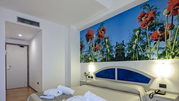 Quarto de hotel com uma cama grande, duas mesas de cabeceira, um quadro com flores vermelhas na parede e uma porta à esquerda.