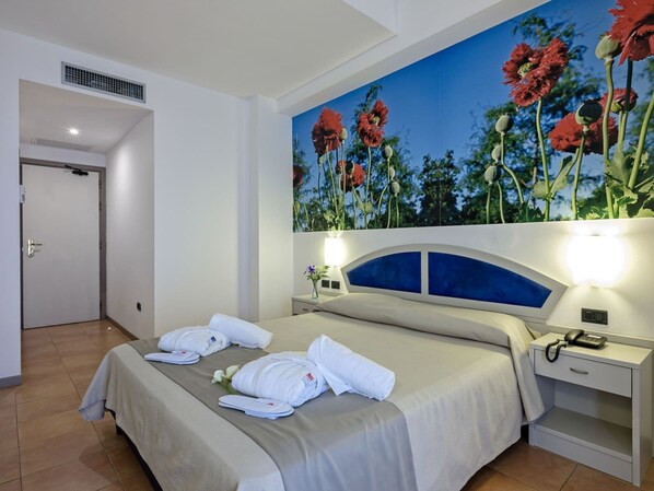 Quarto de hotel com uma cama grande, duas mesas de cabeceira, um quadro com flores vermelhas na parede e uma porta à esquerda.