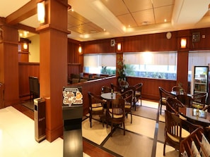 Um saguão de hotel com painéis de madeira, um canto de cafeteria e uma área de estar com mesas e cadeiras.