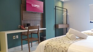 Quarto de hotel com cama, escrivaninha com cadeira, televisão exibindo 'LUMA hotel', espelho e toalha sobre a cama.