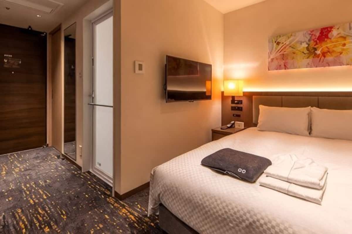 Quarto de hotel com uma cama, televisão fixada na parede, mesa de cabeceira com abajur e um quadro sobre a cama.