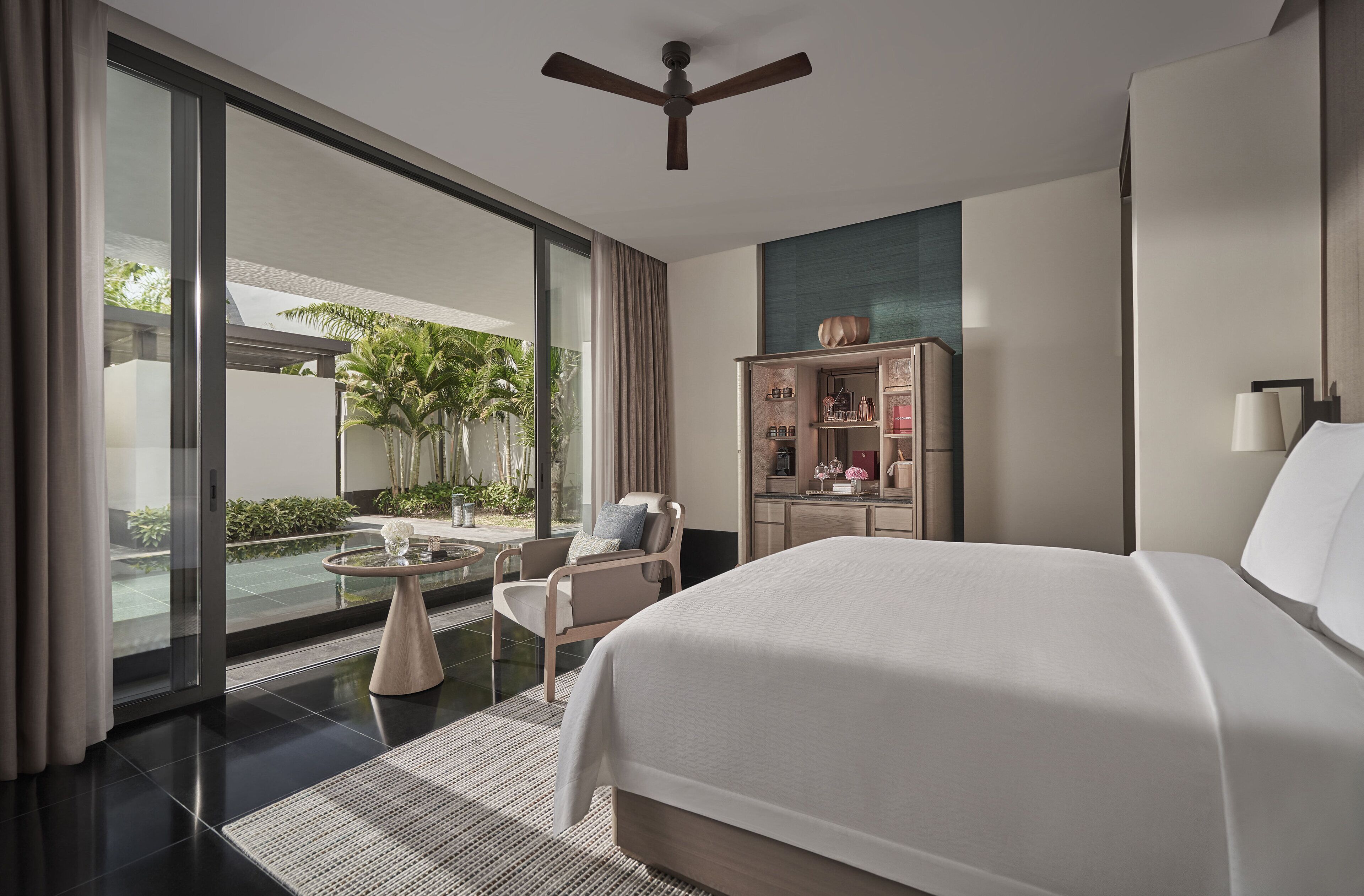 Quarto de hotel moderno com uma cama grande, área de estar com uma mesa, estante de livros e vista para o exterior através de uma porta de vidro.