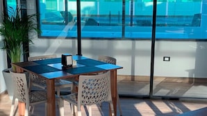 Uma área de refeições com mesa e cadeiras de madeira, grandes janelas com vista para um corpo de água e edifícios, e um vaso de planta.