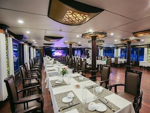 Restaurante com mesas compridas preparadas para refeições, palco ao fundo e vista da cidade à noite.