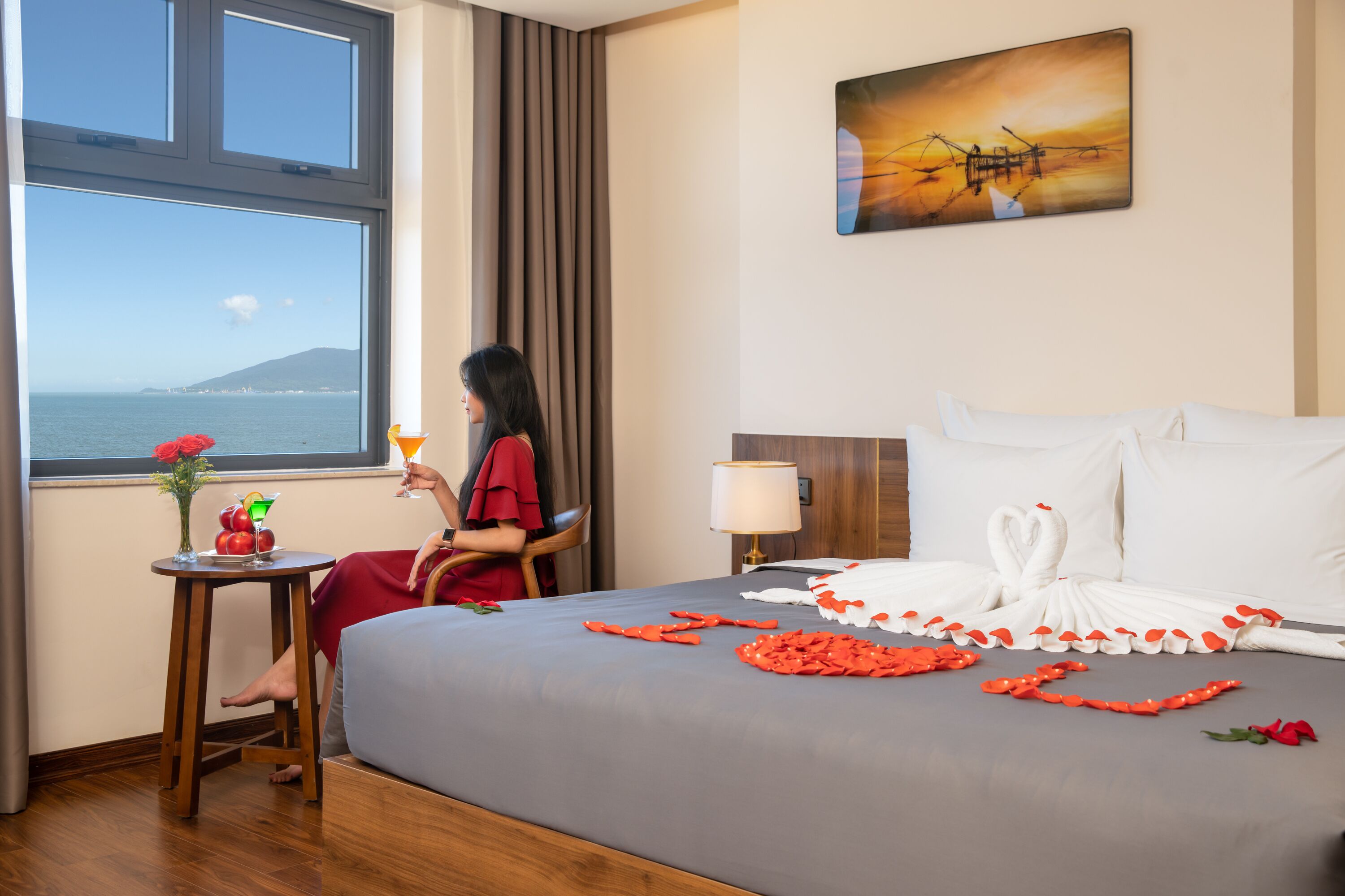 Quarto de hotel com cama, janela vista para o mar, pequena mesa com um vaso de flores e um quadro na parede.