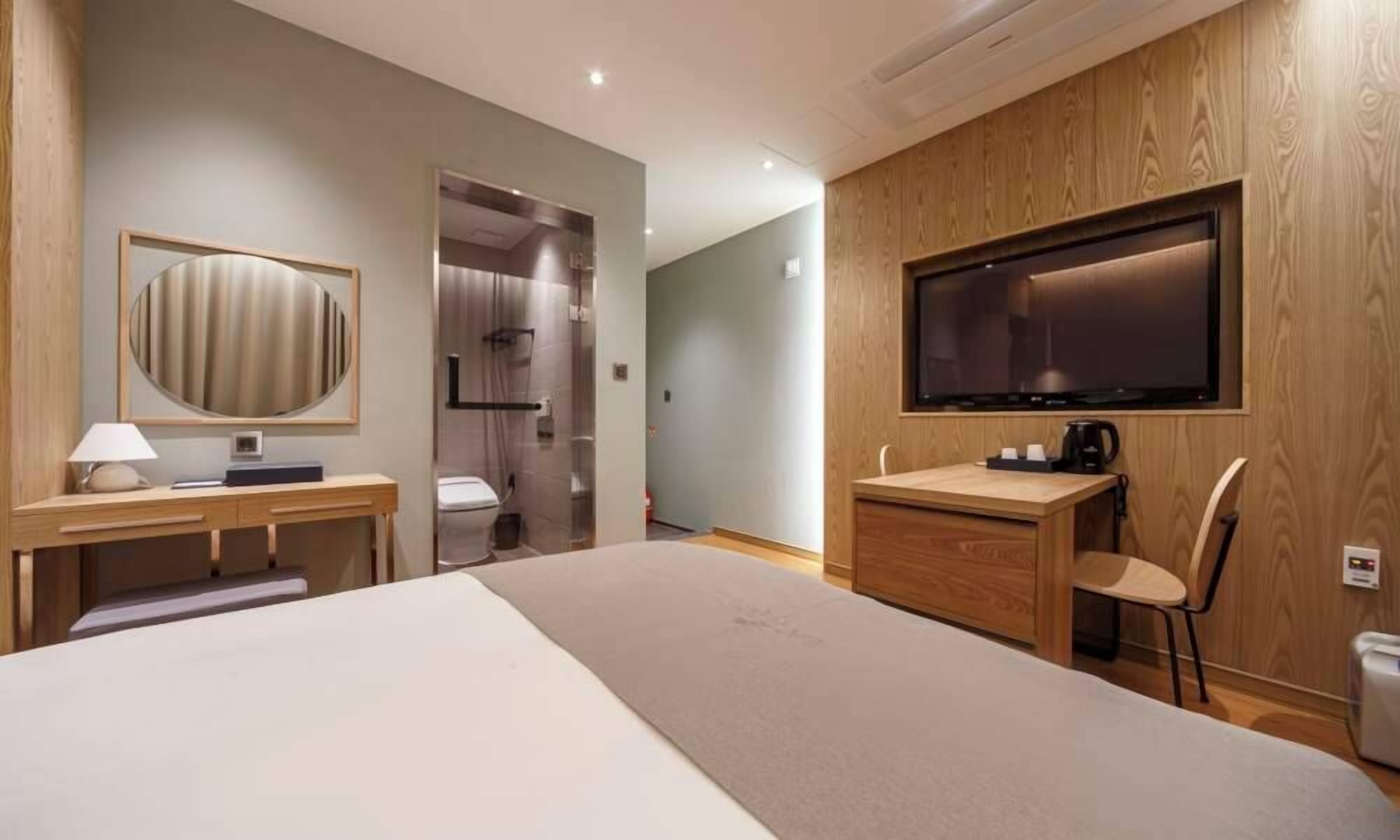 Quarto de hotel com cama, escrivaninha com abajur, televisão e área de chuveiro.