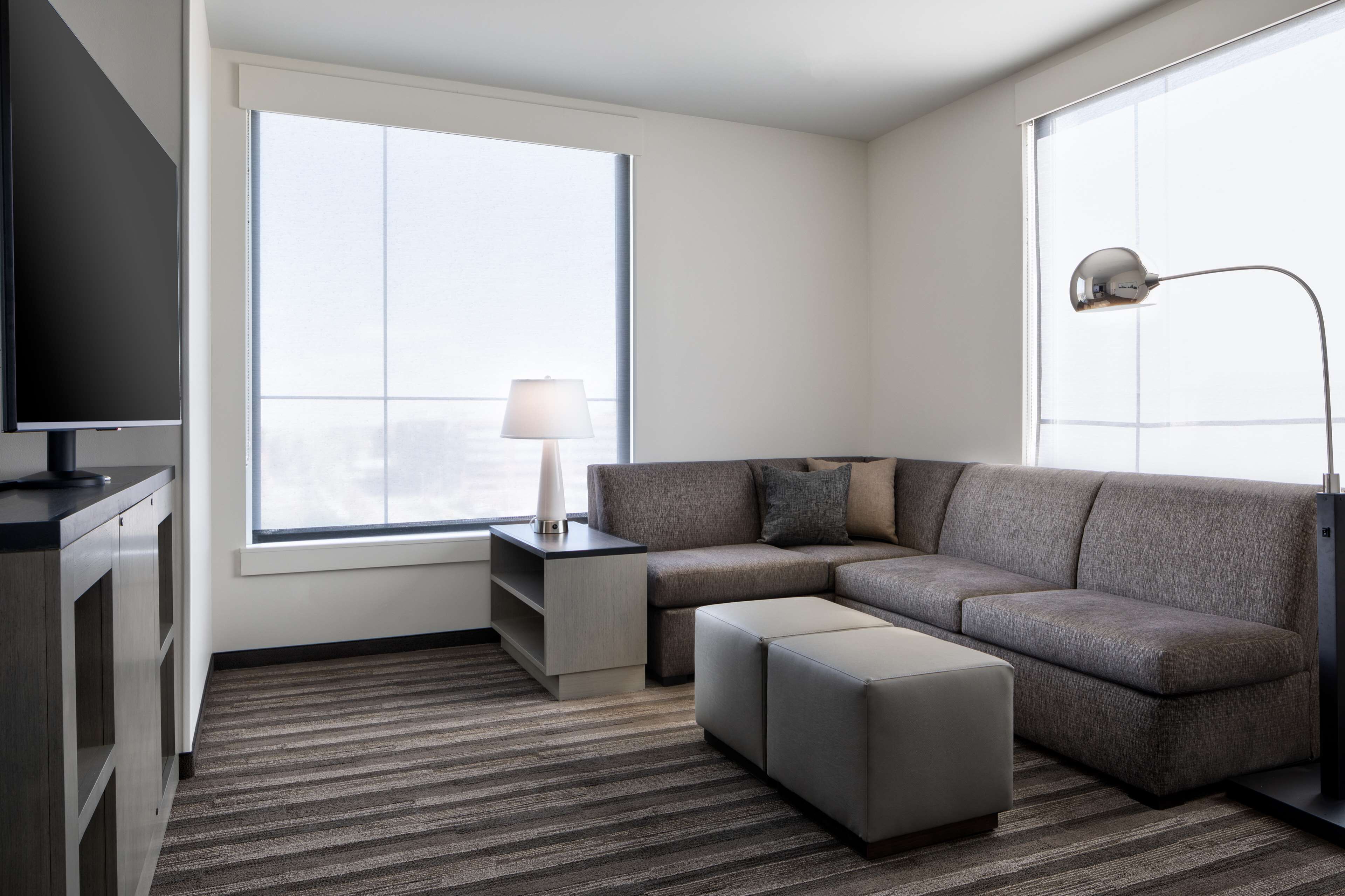 Quarto de hotel moderno com sofá modular, TV de tela plana, abajur e uma grande janela com vista.