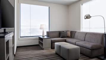 Quarto de hotel moderno com sofá modular, TV de tela plana, abajur e uma grande janela com vista.