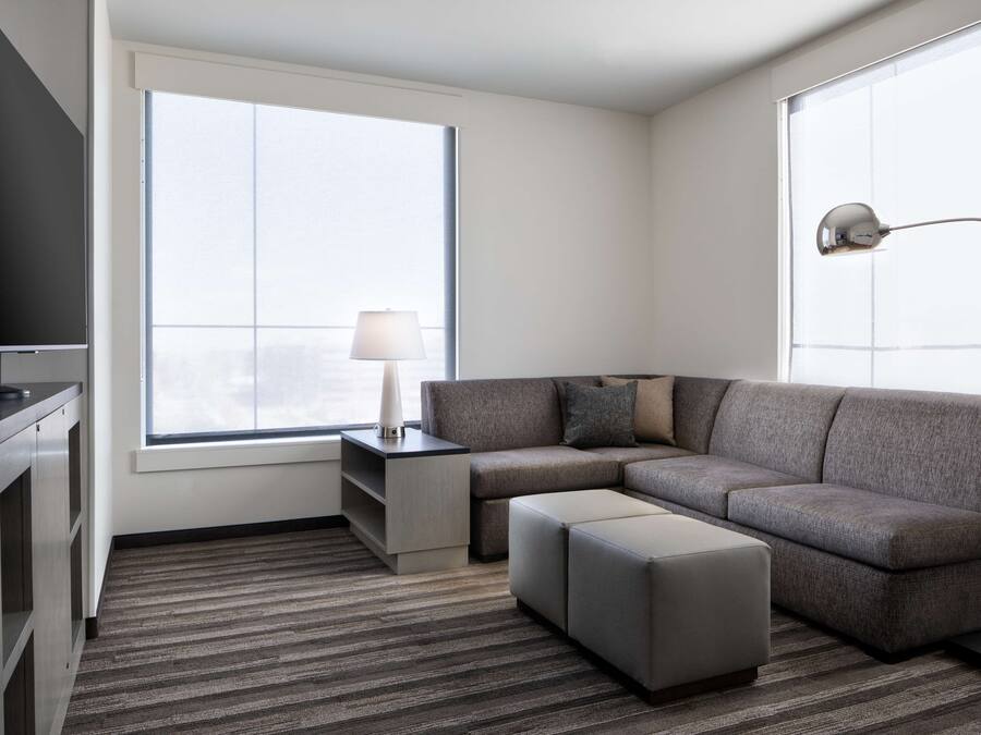 Quarto de hotel moderno com sofá modular, TV de tela plana, abajur e uma grande janela com vista.