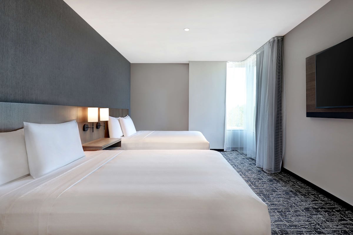 Quarto de hotel com duas camas, televisão de tela plana, mesa de cabeceira com abajur e uma janela grande com cortinas.