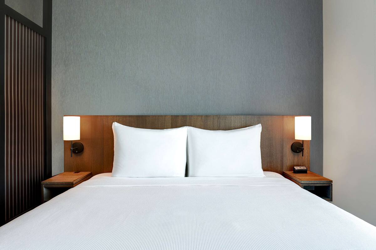 Uma cama bem arrumada com roupa de cama branca, duas mesas de cabeceira com luminárias e uma cabeceira de madeira.