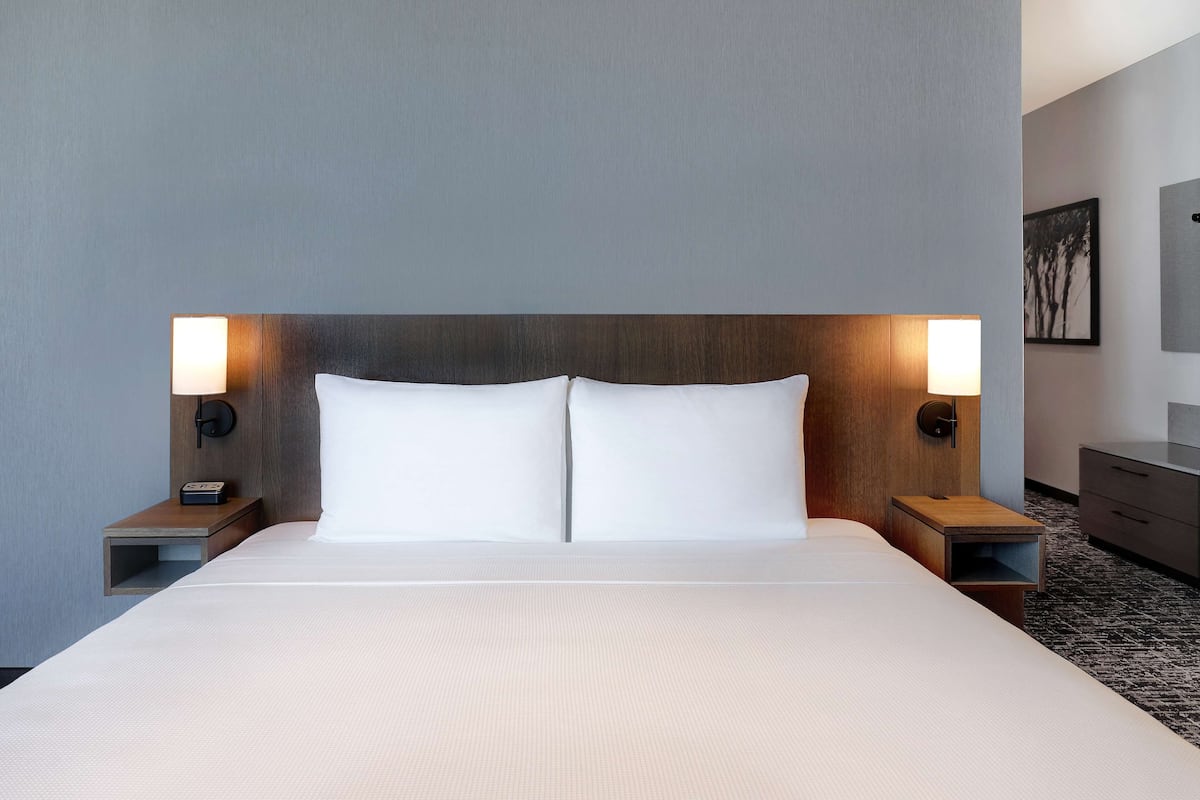 Quarto de hotel moderno com uma cama grande, duas mesas de cabeceira com luminárias, uma cômoda e um quadro na parede.