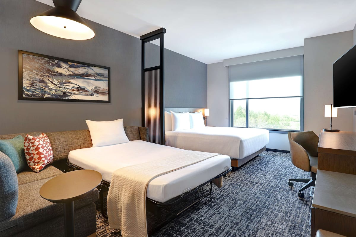 Quarto de hotel com duas camas, um sofá, uma mesinha, uma escrivaninha e uma janela grande.