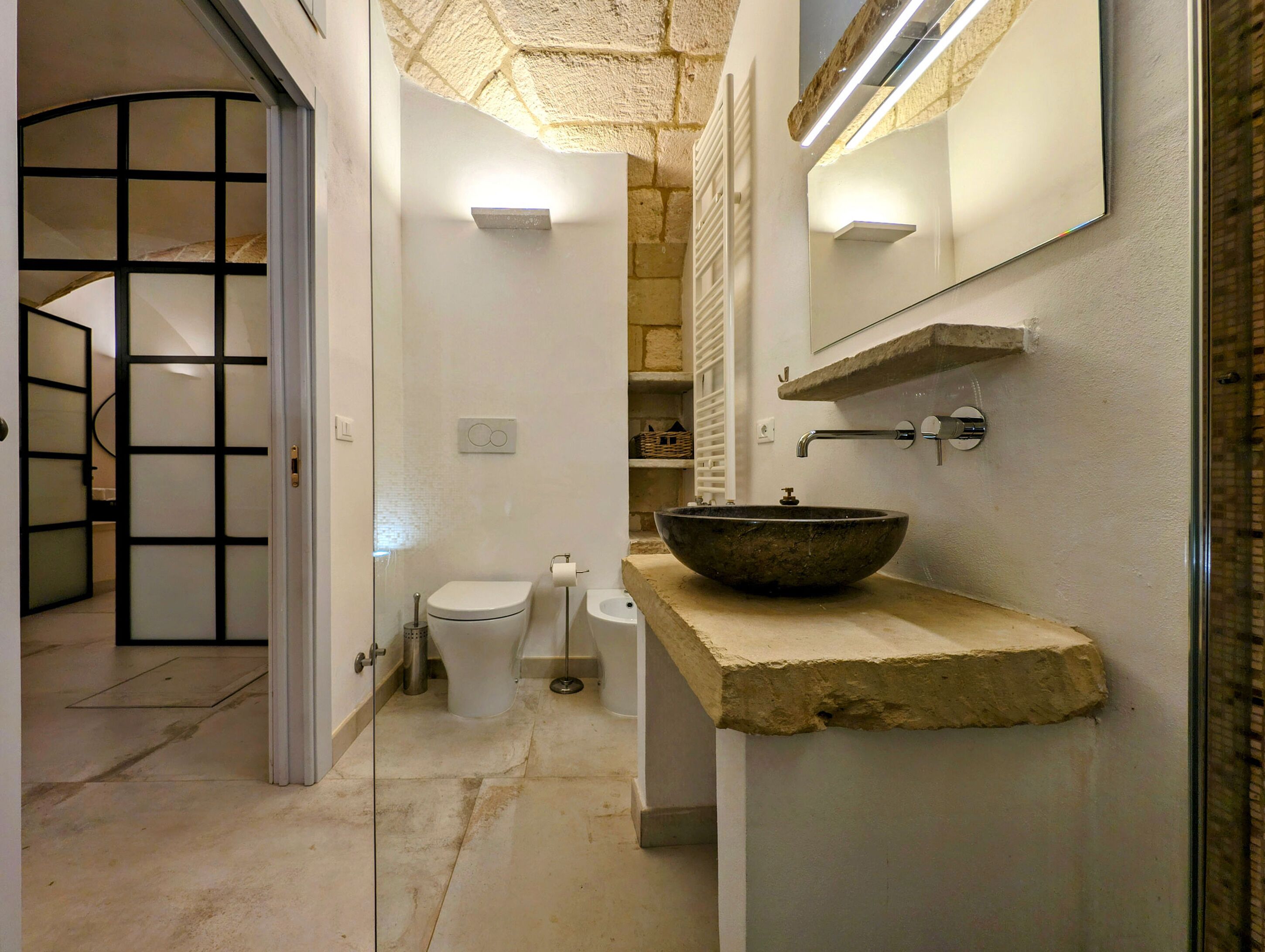 Casa de banho com lavatório de pedra, sanita branca e porta de vidro para outro espaço.