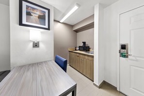Quarto de hotel com mesa de jantar, uma cadeira azul, cozinha compacta e porta com fechadura eletrônica.