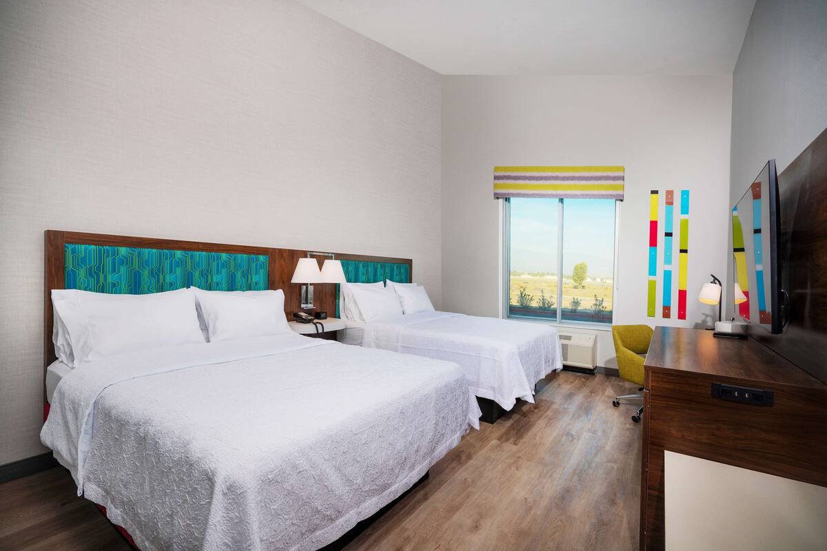 Quarto de hotel com duas camas, uma escrivaninha, uma cadeira, uma televisão e uma janela com vista.