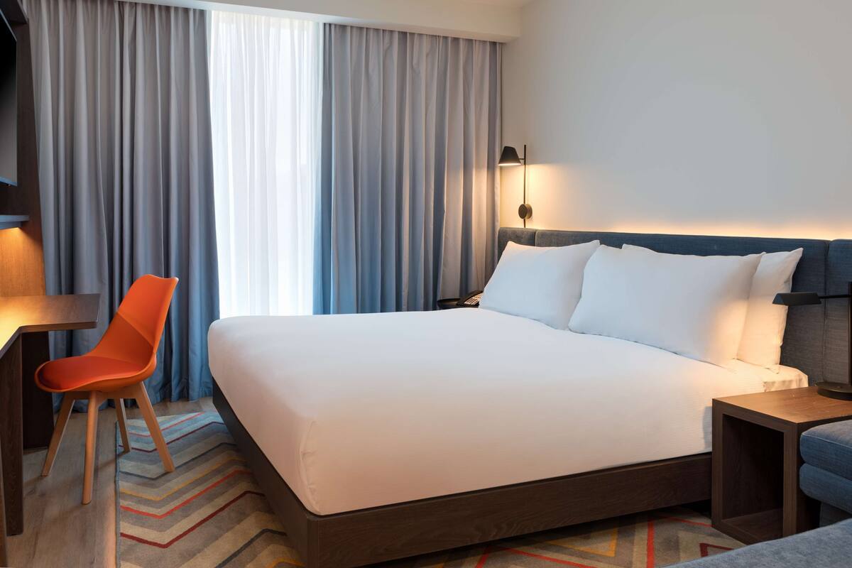 Quarto de hotel com uma cama grande, uma mesa, uma cadeira laranja e uma mesa de cabeceira com abajur.