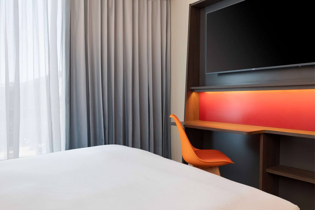 Quarto de hotel com cama, televisão fixada na parede, escrivaninha com luz vermelha e uma cadeira.