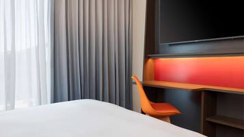 Quarto de hotel com cama, televisão fixada na parede, escrivaninha com luz vermelha e uma cadeira.