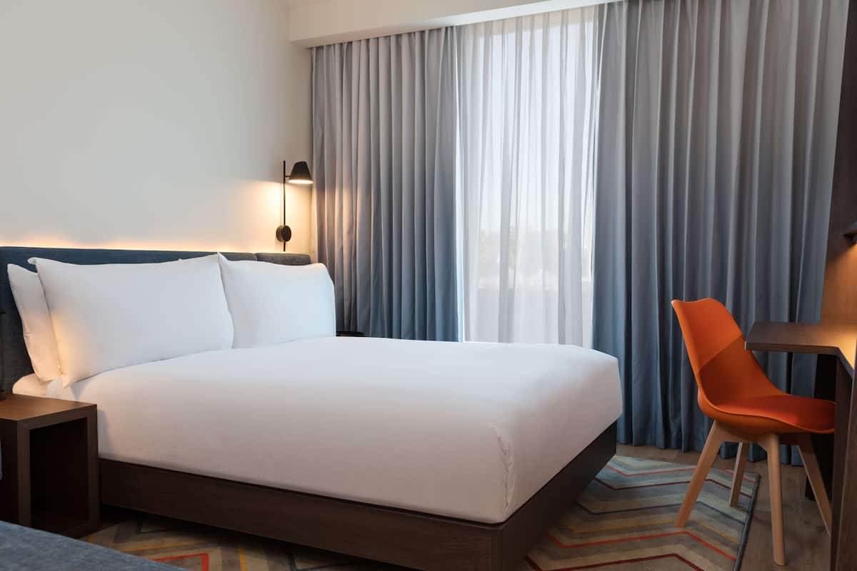 Quarto de hotel com uma cama grande, uma escrivaninha com cadeira, uma luminária e vista através de cortinas leves.