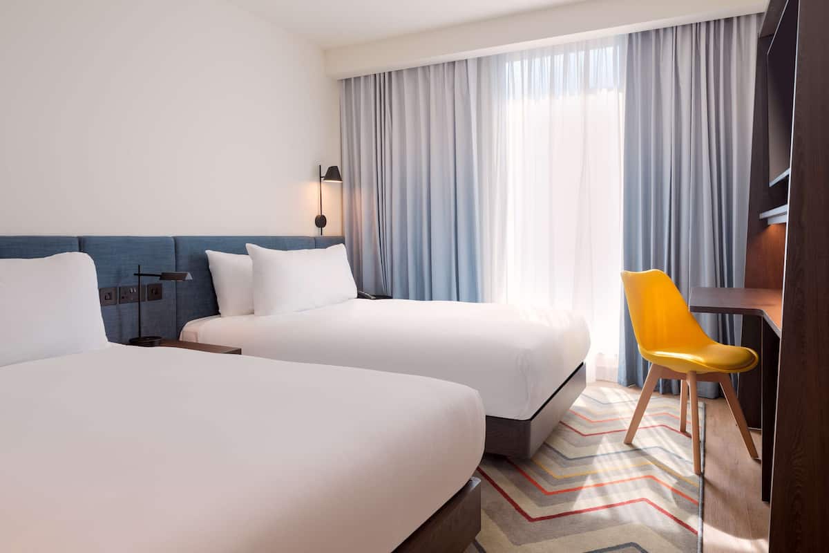 Quarto de hotel com duas camas, uma escrivaninha, uma cadeira e uma janela grande com cortinas.