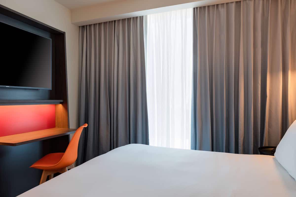 Quarto de hotel com cama, escrivaninha com cadeira, televisão e cortinas grandes.