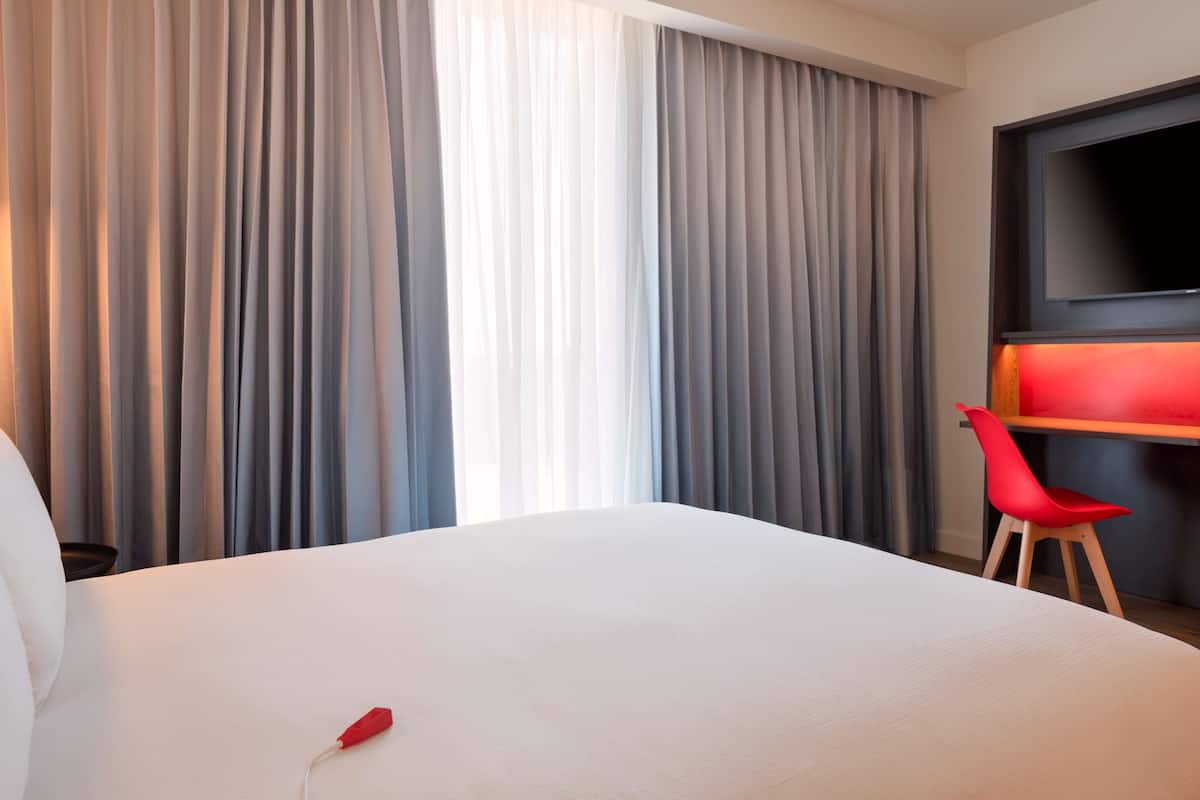 Quarto de hotel com uma cama grande, uma escrivaninha e uma cadeira vermelha, e uma televisão na parede.