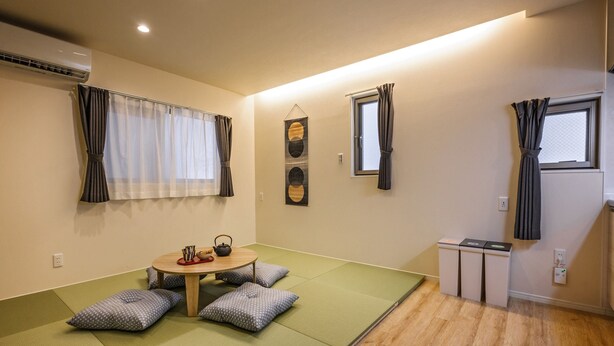 Quarto com esteiras de tatame, uma mesa redonda com almofadas, uma pequena prateleira com objetos e uma janela com cortinas.