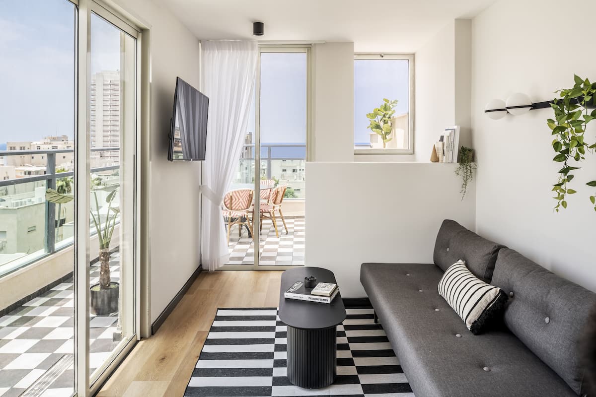 Sala de estar moderna com sofá cinza, mesa de centro preta e tapete xadrez. Possui uma varanda com vista para a cidade e uma planta no parapeito da janela.