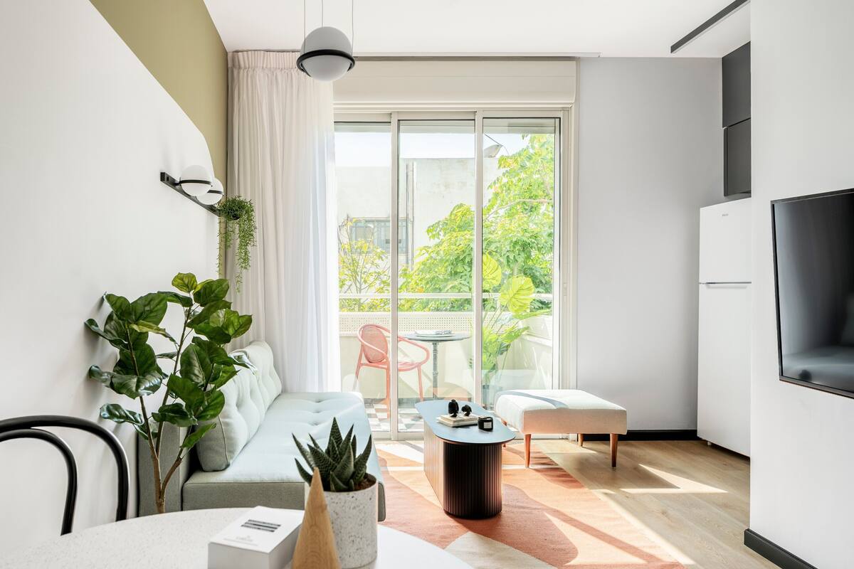 Uma sala de estar moderna com um sofá, uma mesa de centro e uma TV. Há uma varanda com uma mesa e cadeiras, e uma janela grande que oferece vista para o verde.