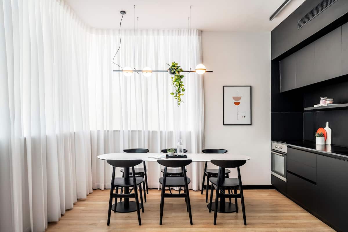 Uma cozinha moderna com bancada preta e armários brancos, uma área de jantar com mesa branca e cadeiras pretas, e uma janela grande com cortinas brancas.