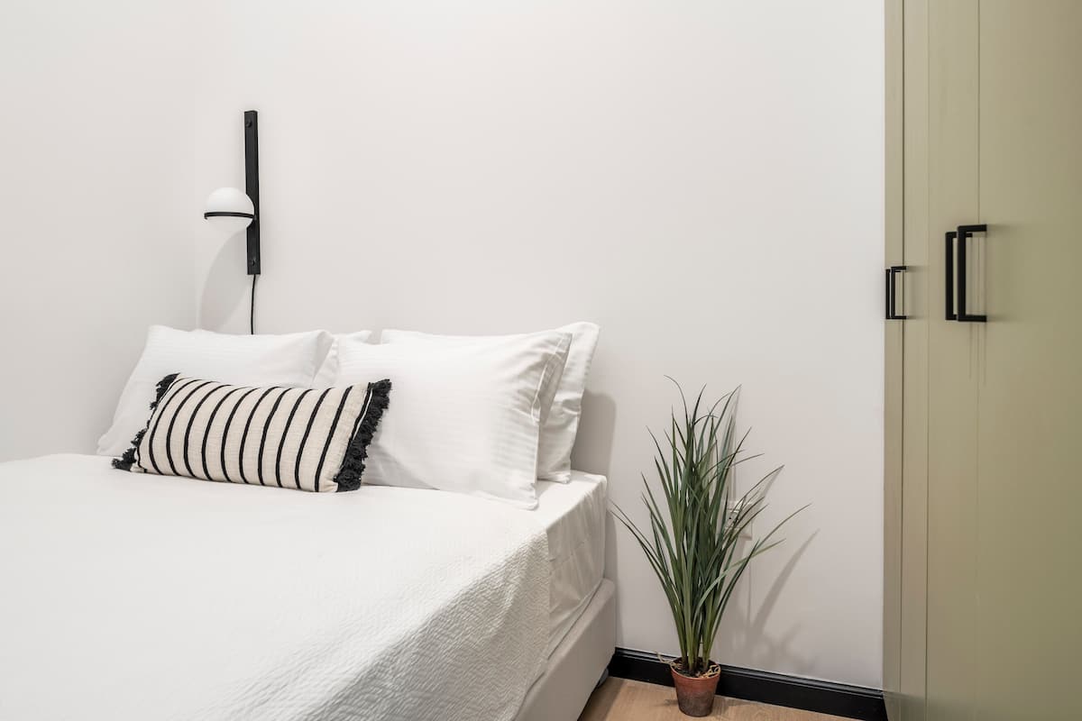 Cama bem arrumada com roupa de cama branca e almofadas listradas, uma planta em vaso e uma luminária fixada na parede.