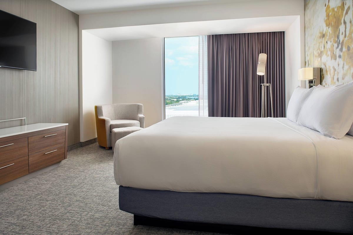 Quarto de hotel com cama grande, televisão de tela plana na parede, uma cadeira, escrivaninha com gavetas e uma janela ampla com vista para a cidade.