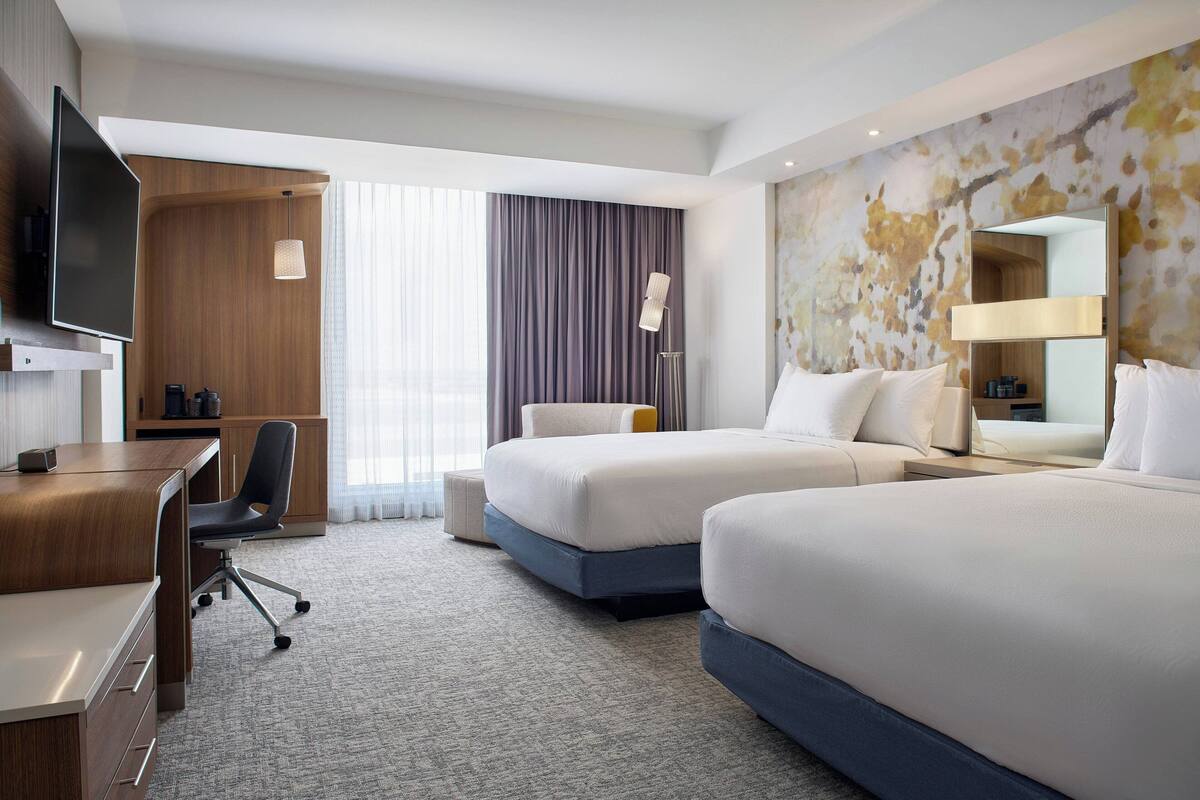 Quarto de hotel com duas camas, uma mesa, uma cadeira, televisão e uma janela grande com cortinas.