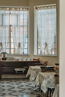 Uma área de refeições com duas janelas, uma cafeteira, um rádio e uma mesa posta com pratos e chávenas.