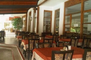 Restaurante com várias mesas e cadeiras, janelas amplas e uma planta ao fundo.