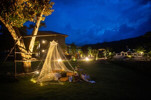 Uma tenda montada numa área gramada, com luzes de corda, uma mesa com uma garrafa e copos, e uma árvore por perto.
