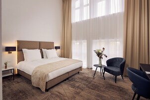 Quarto de hotel com uma cama grande, uma mesa pequena com abajur, uma cadeira e vista para o exterior através de cortinas leves.