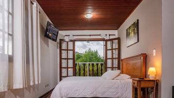 Quarto com teto de madeira, cama com roupa de cama branca, mesa de cabeceira com abajur, janela com cortinas e vista para o verde lá fora.