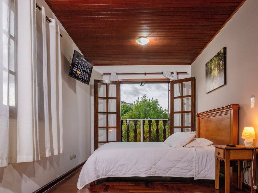 Quarto com teto de madeira, cama com roupa de cama branca, mesa de cabeceira com abajur, janela com cortinas e vista para o verde lá fora.