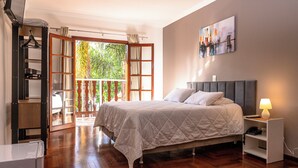 Quarto com cama, mesa de cabeceira, pintura e vista para o verde através de uma porta aberta.