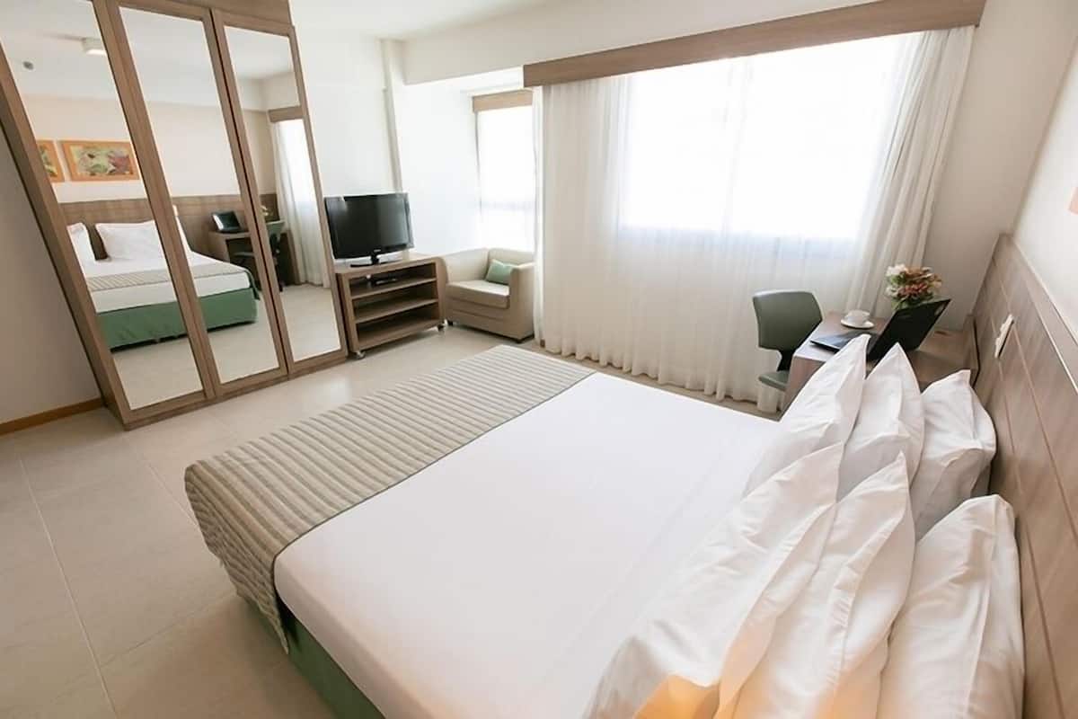 Quarto de hotel com uma cama grande, televisão, escrivaninha com cadeira e espelho.