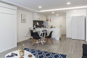 Uma cozinha moderna com armários brancos, um espaço de jantar com mesa redonda e cadeiras pretas, e uma pequena geladeira.