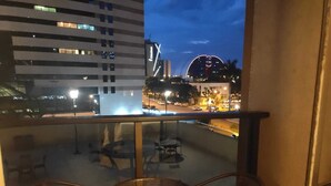 Varanda com cadeiras de vime e mesa de vidro, com vista para uma rua com edifícios e uma roda gigante.