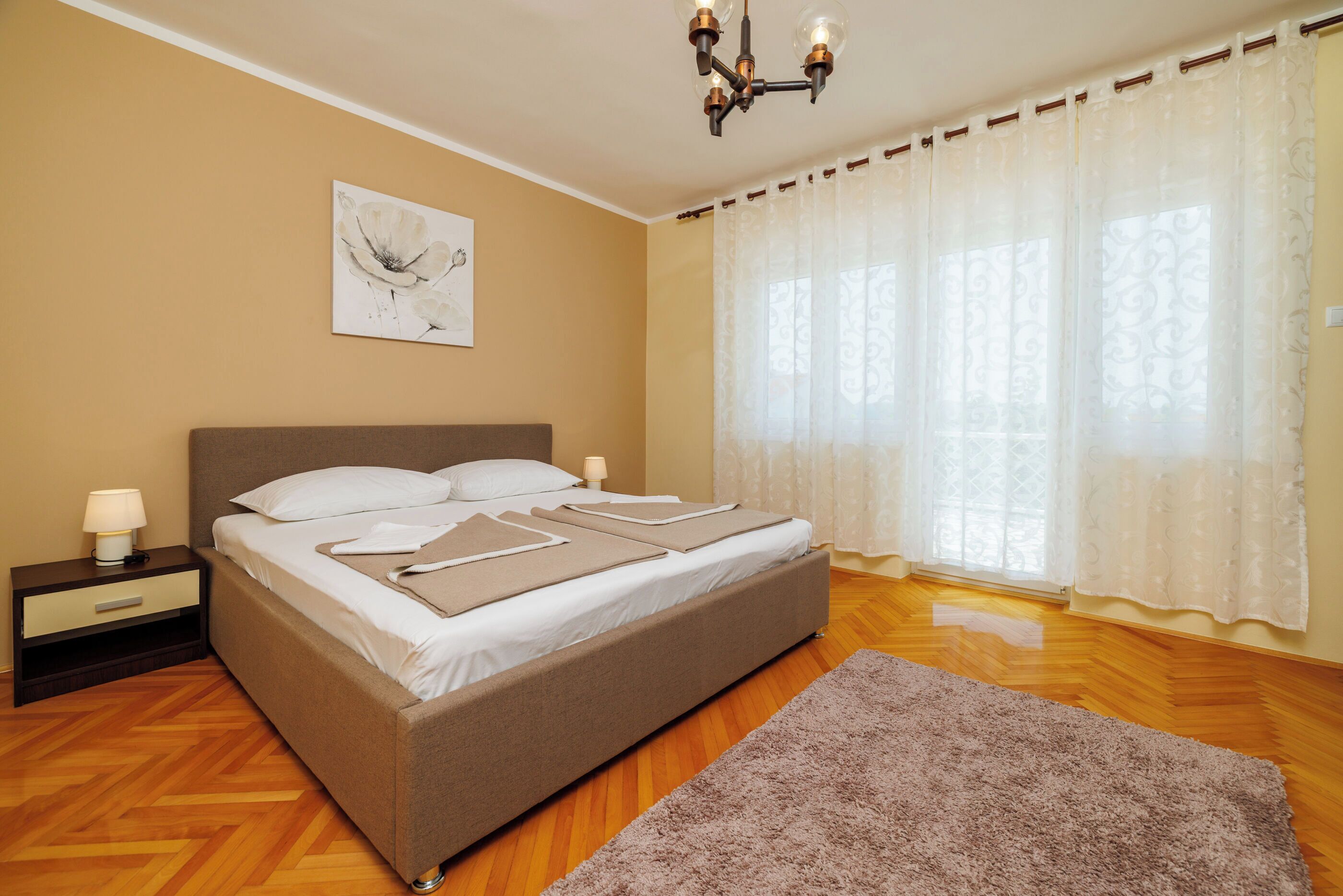 Um quarto com cama, mesa de cabeceira, luminária, pintura e janela com cortinas.