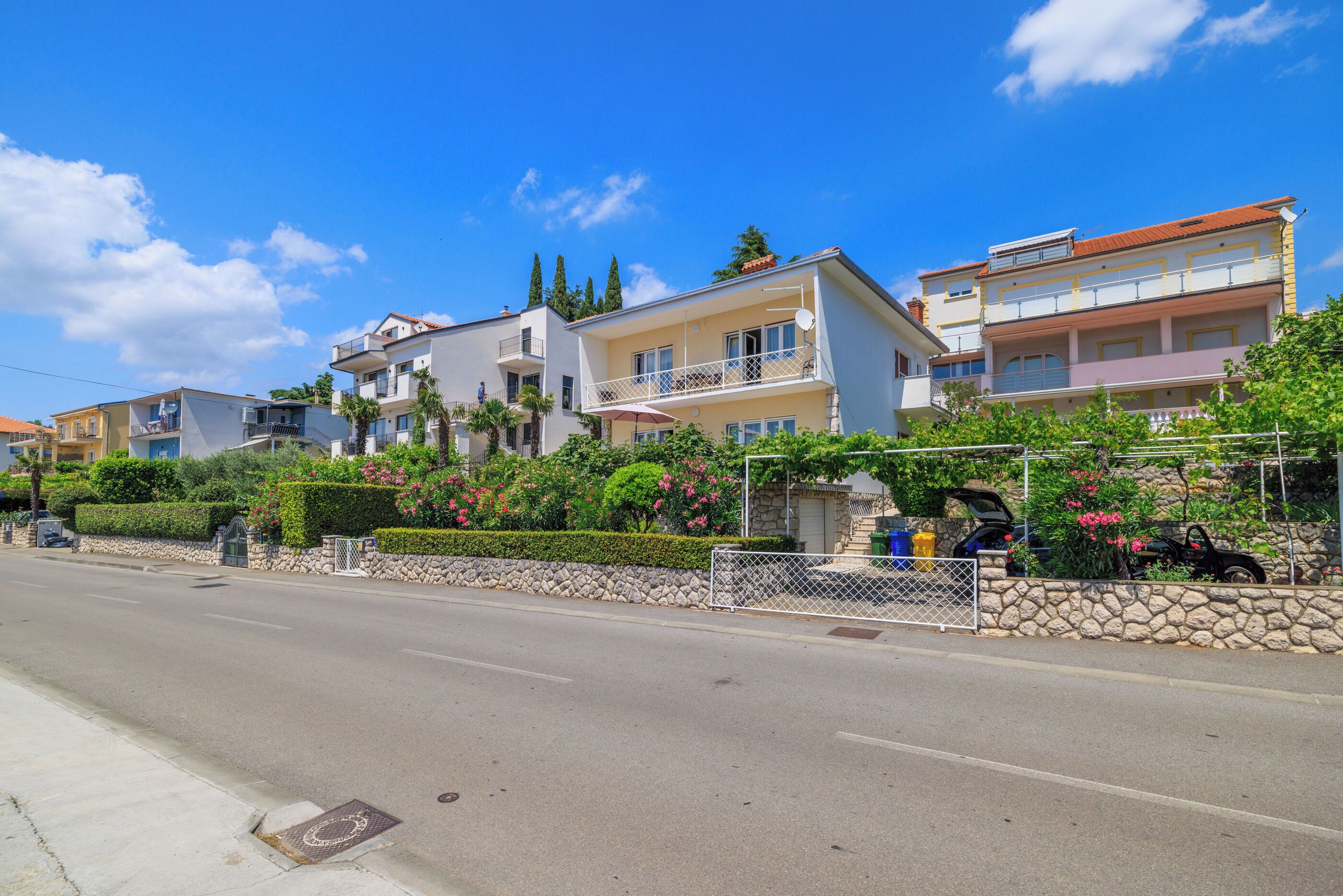 Rua residencial com casas de vários andares, uma parede de pedra e um céu azul límpido.