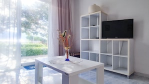 Uma sala de estar moderna com uma mesa branca, um vaso com flores secas, uma televisão sobre um suporte branco e vista para o verde lá fora.