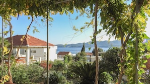 Vista costeira com um edifício branco, telhado de telhas vermelhas e um jardim com diversas plantas e um banco.