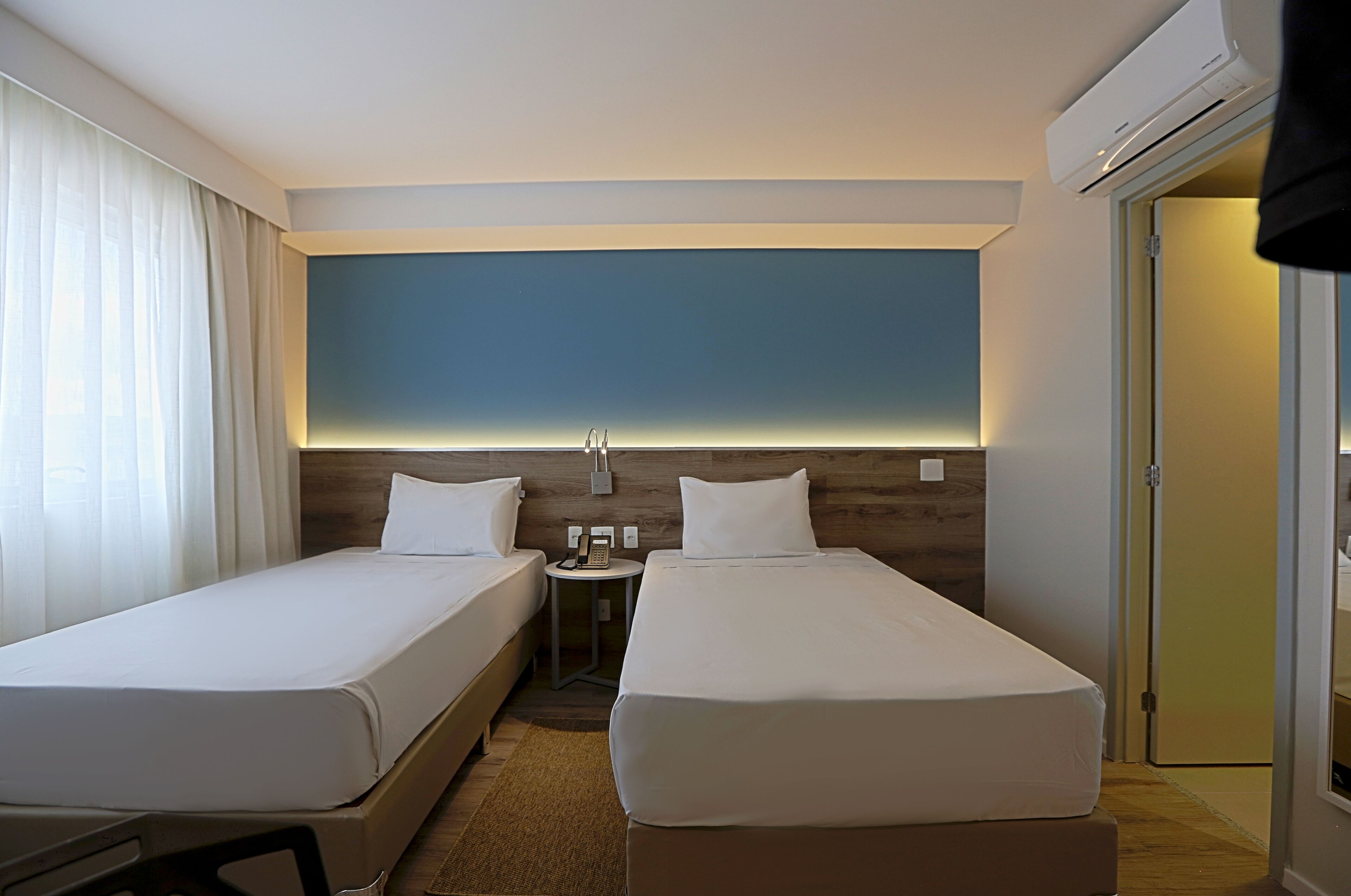 Quarto de hotel com duas camas de solteiro, mesa de cabeceira com abajur e ar-condicionado na parede.