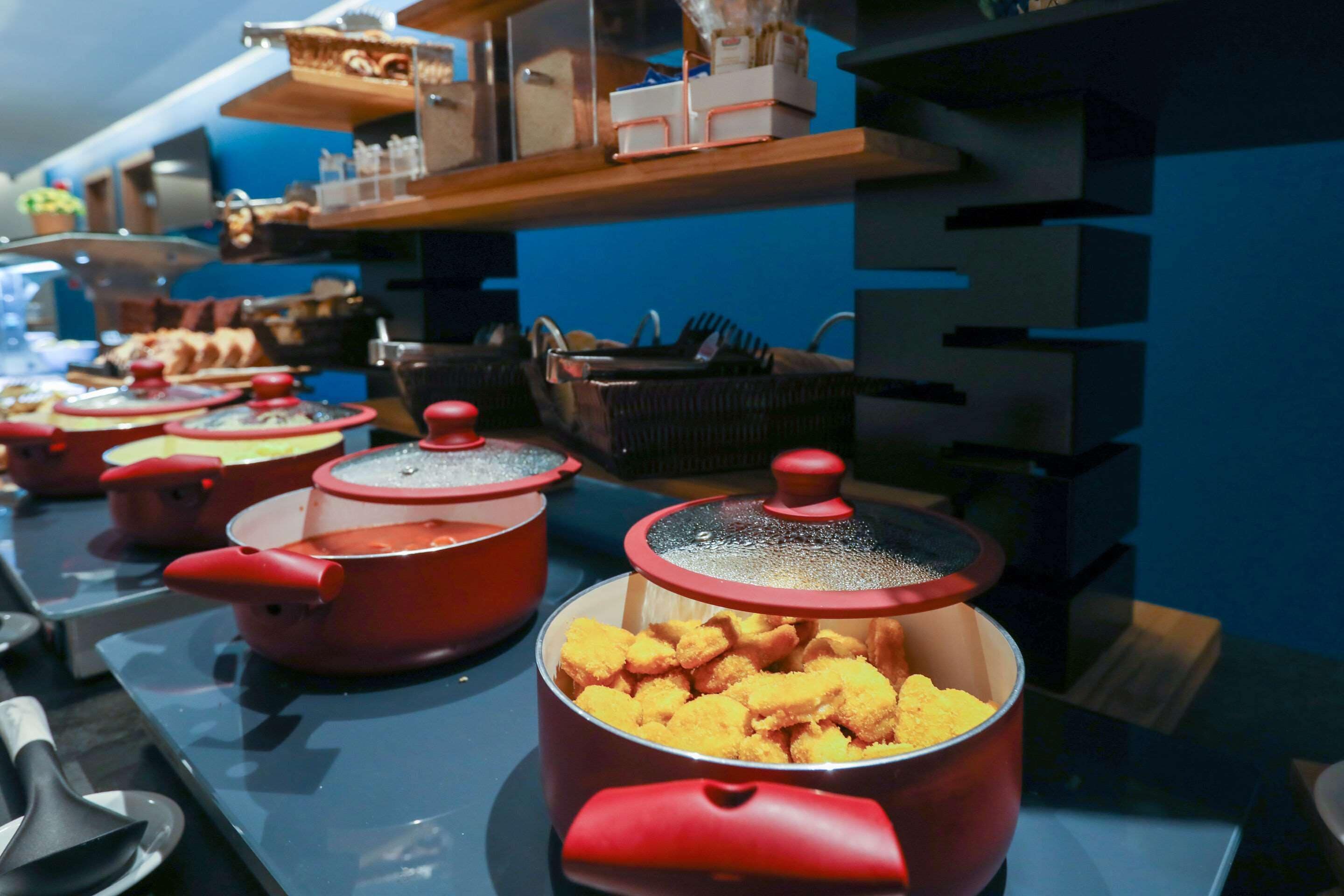 Disposição de buffet com diversos alimentos em panelas vermelhas, parede azul e uma prateleira com utensílios de cozinha.